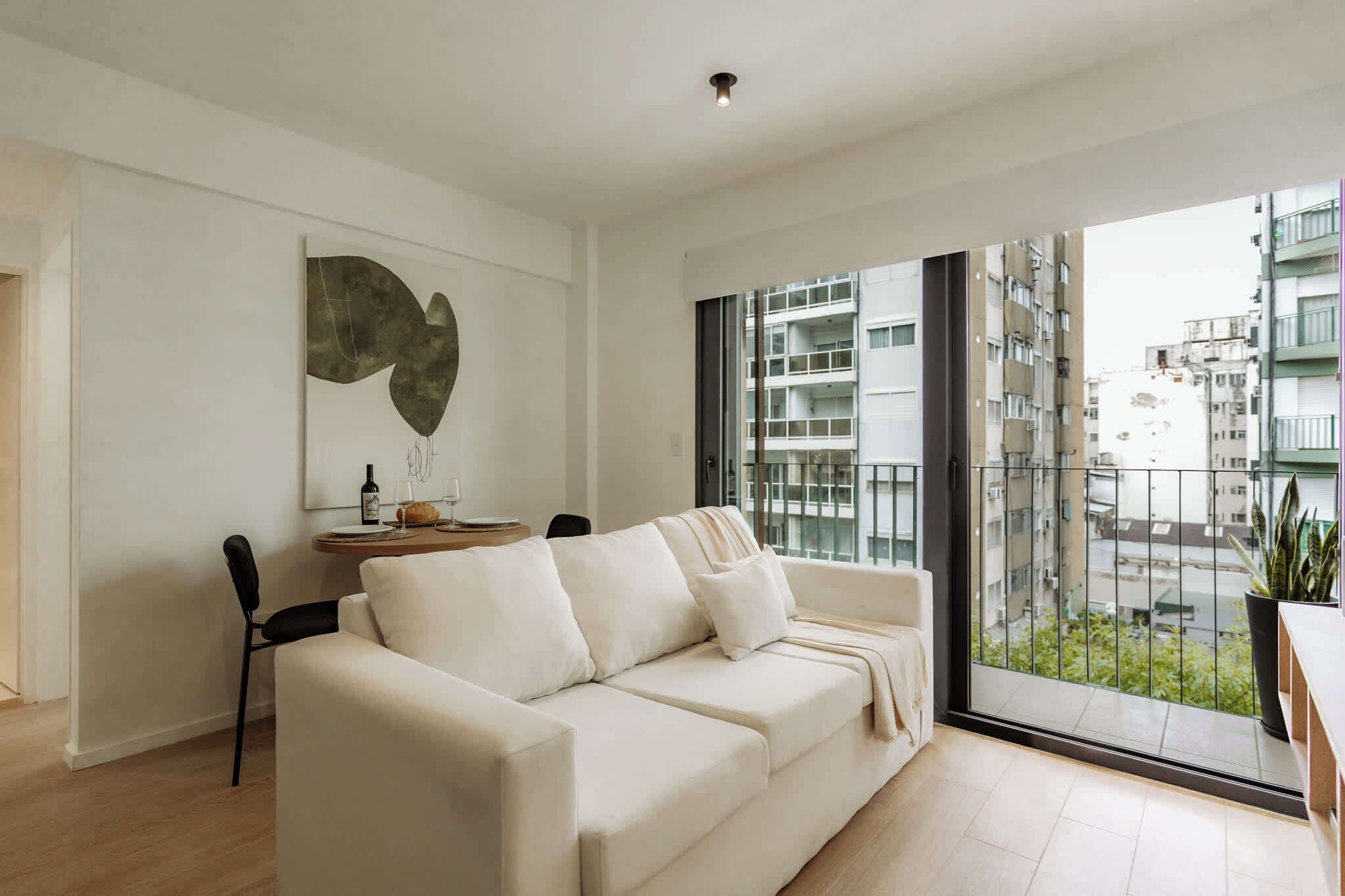 Uma sala de estar moderna com um sofá branco, uma mesa de jantar com garrafa e copos, e uma grande janela com vista para edifícios.