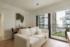 Uma sala de estar moderna com um sofá branco, uma mesa de jantar com garrafa e copos, e uma grande janela com vista para edifícios.