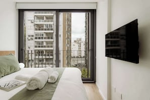 Quarto de hotel com uma cama, um cobertor verde, duas toalhas enroladas, uma janela com vista para edifícios e uma televisão na parede.