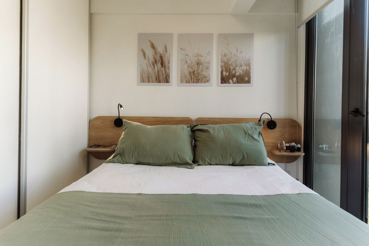 Quarto moderno com cama grande, roupa de cama verde, cabeceira de madeira, mesas de cabeceira com luminárias e três quadros pendurados na parede.