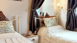 Quarto com duas camas, cortinas escuras, mesa de cabeceira com relógio, criado-mudo e luminária na parede.