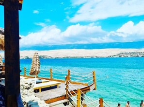 Um píer de madeira com um toldo de palha, que se estende até um mar azul e límpido, com uma praia de areia branca ao longe e um céu claro.