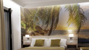Quarto com cama, mesas de cabeceira, abajur e um mural de palmeiras e pôr do sol.