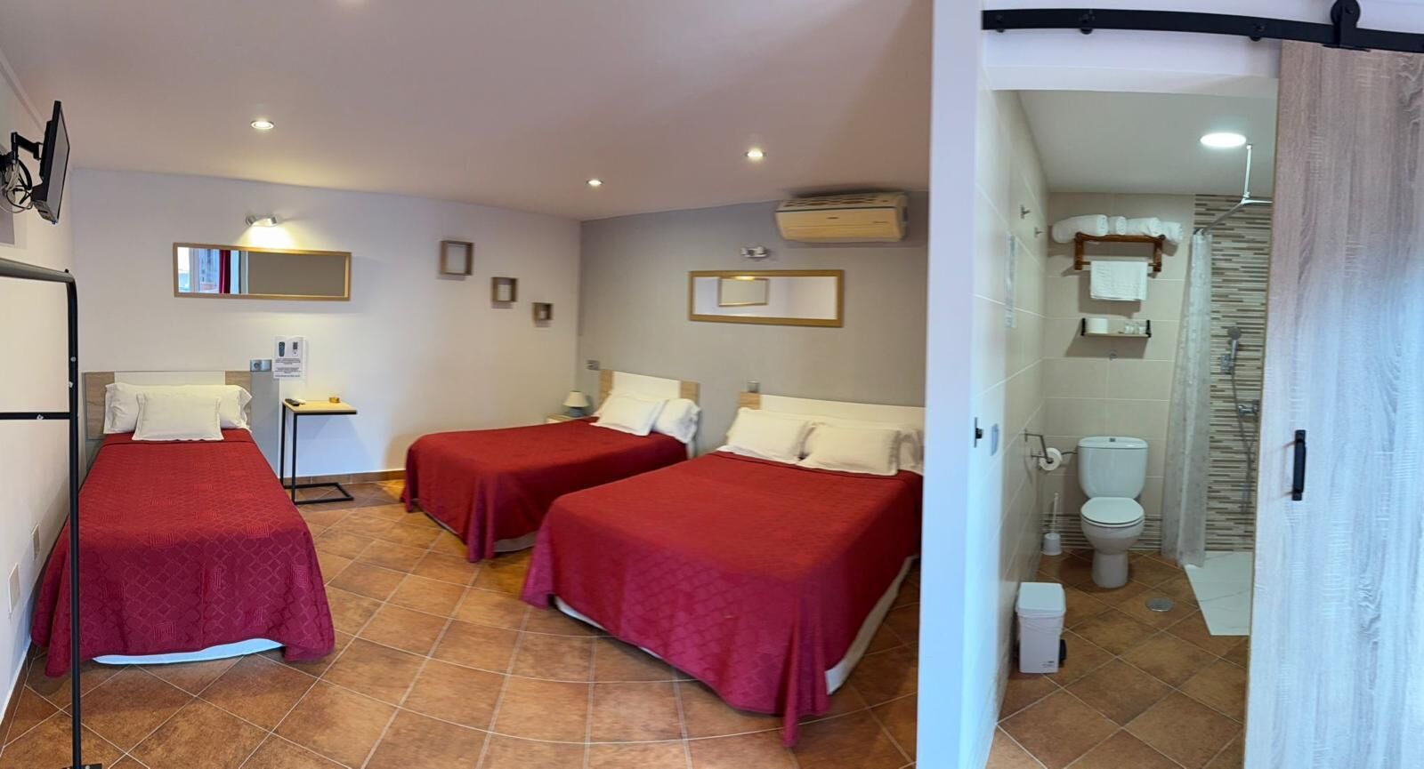 Quarto de hotel com duas camas, banheiro com chuveiro e uma pequena mesa com luminária.