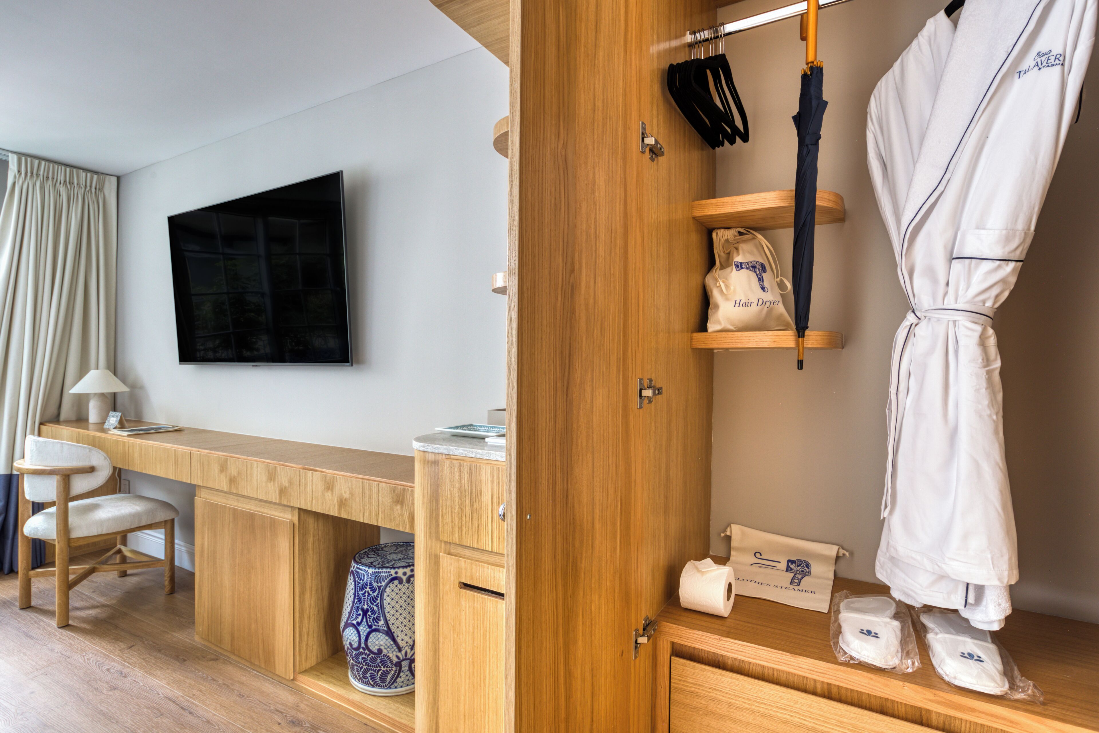 Quarto de hotel com secretária de madeira, uma cadeira, televisão, guarda-roupa e área de banheiro com toalha e secador de cabelo.