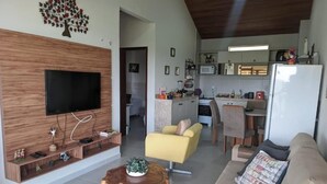Sala de estar com televisão, móvel de madeira para TV, sofá amarelo e um espaço de jantar com cadeiras e mesa.