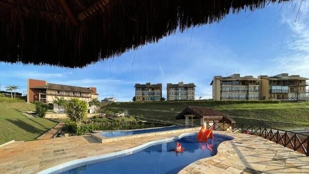 Uma piscina com escorrega, rodeada por um deck de pedra, e edifícios ao fundo.