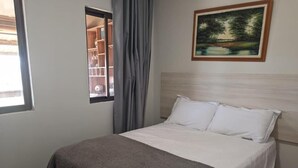 Quarto com cama, janela com cortinas, quadro na parede e vista para um cômodo com prateleiras.