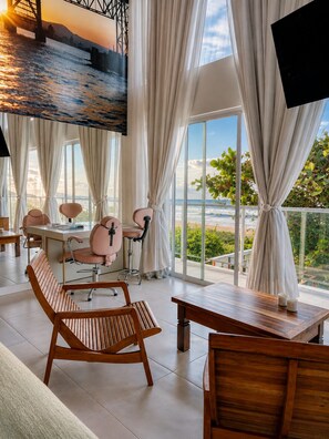 Um quarto amplo com uma grande janela com vista para uma praia, mobiliado com cadeiras e uma mesa de madeira.