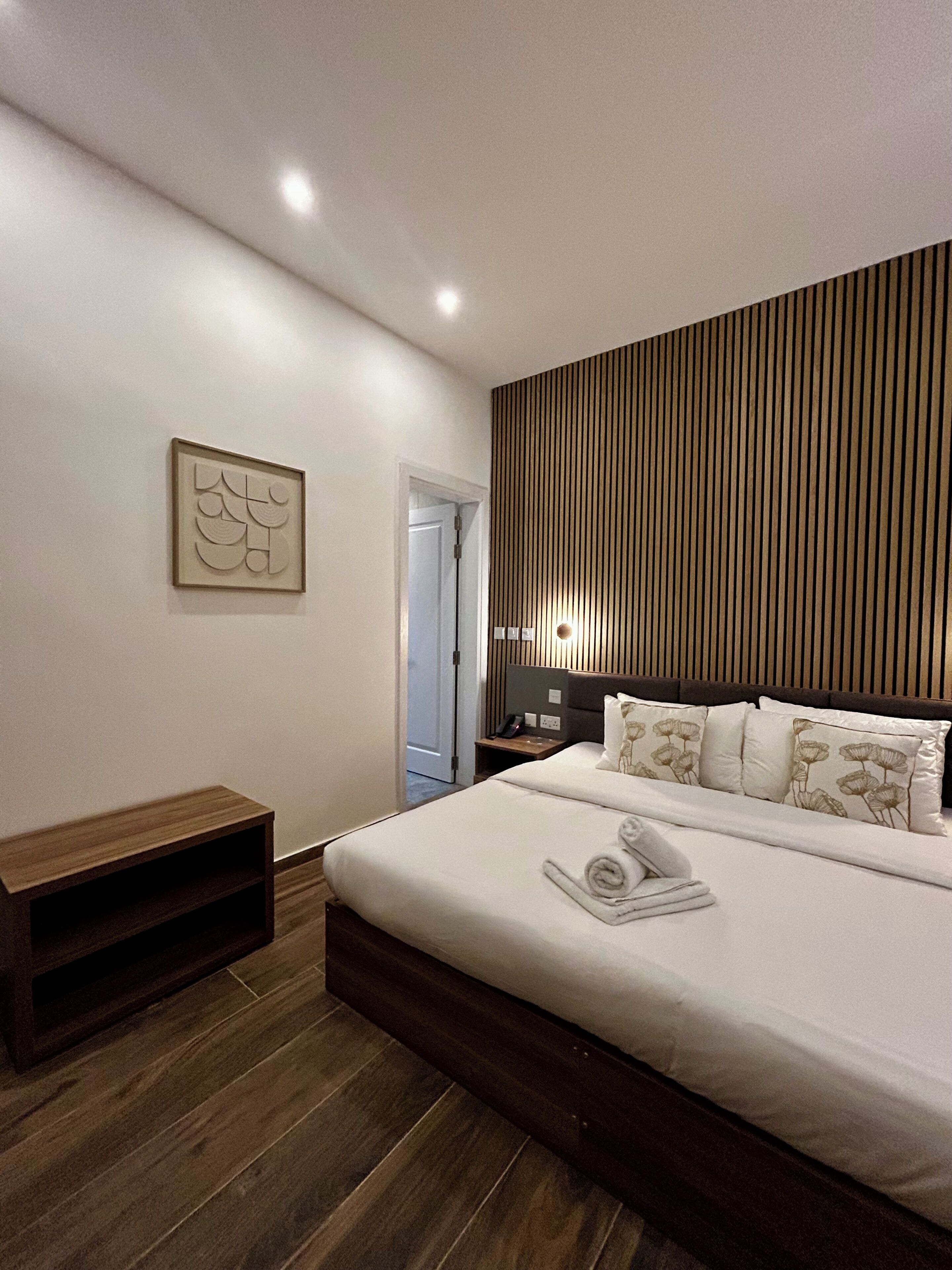 Um quarto moderno com uma cama grande, uma mesa de cabeceira, um banco de madeira, uma luminária fixada na parede e uma obra de arte emoldurada.