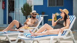 Duas pessoas relaxando em espreguiçadeiras junto a uma piscina, com um edifício azul e um extintor de incêndio ao fundo.