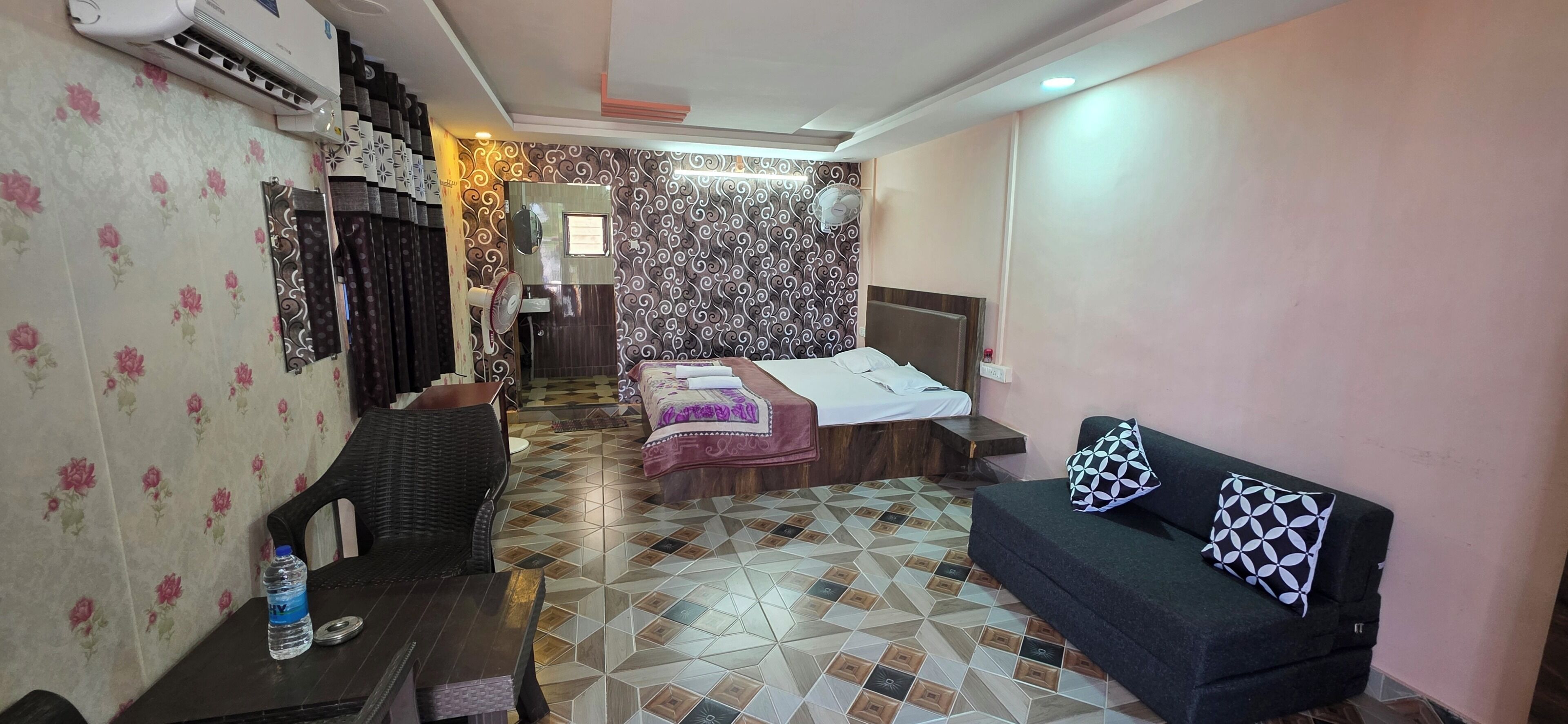 Quarto de hotel com cama, sofá, mesa, garrafa de água, ventilador e ar condicionado fixado na parede.