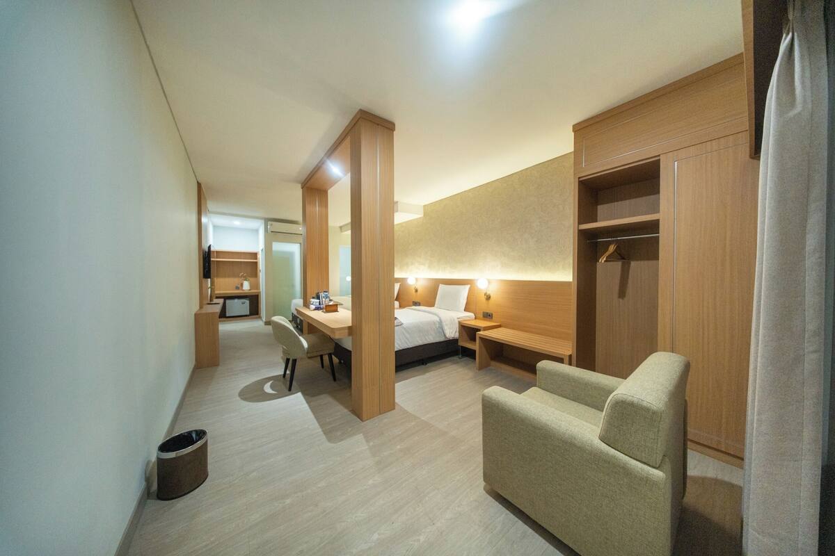Quarto de hotel com cama, escrivaninha, cadeira, guarda-roupa e um pequeno cesto de lixo.