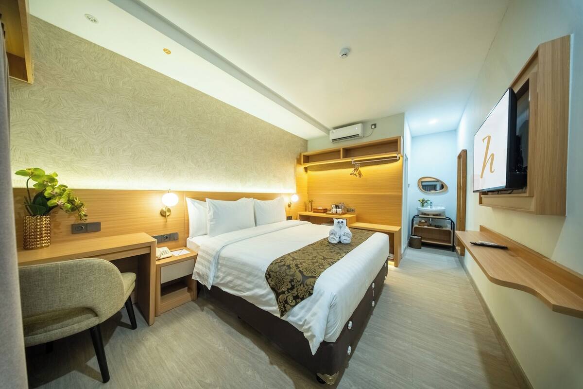 Quarto de hotel com cama, mesa de cabeceira, luminária, televisão e escrivaninha com cadeira.