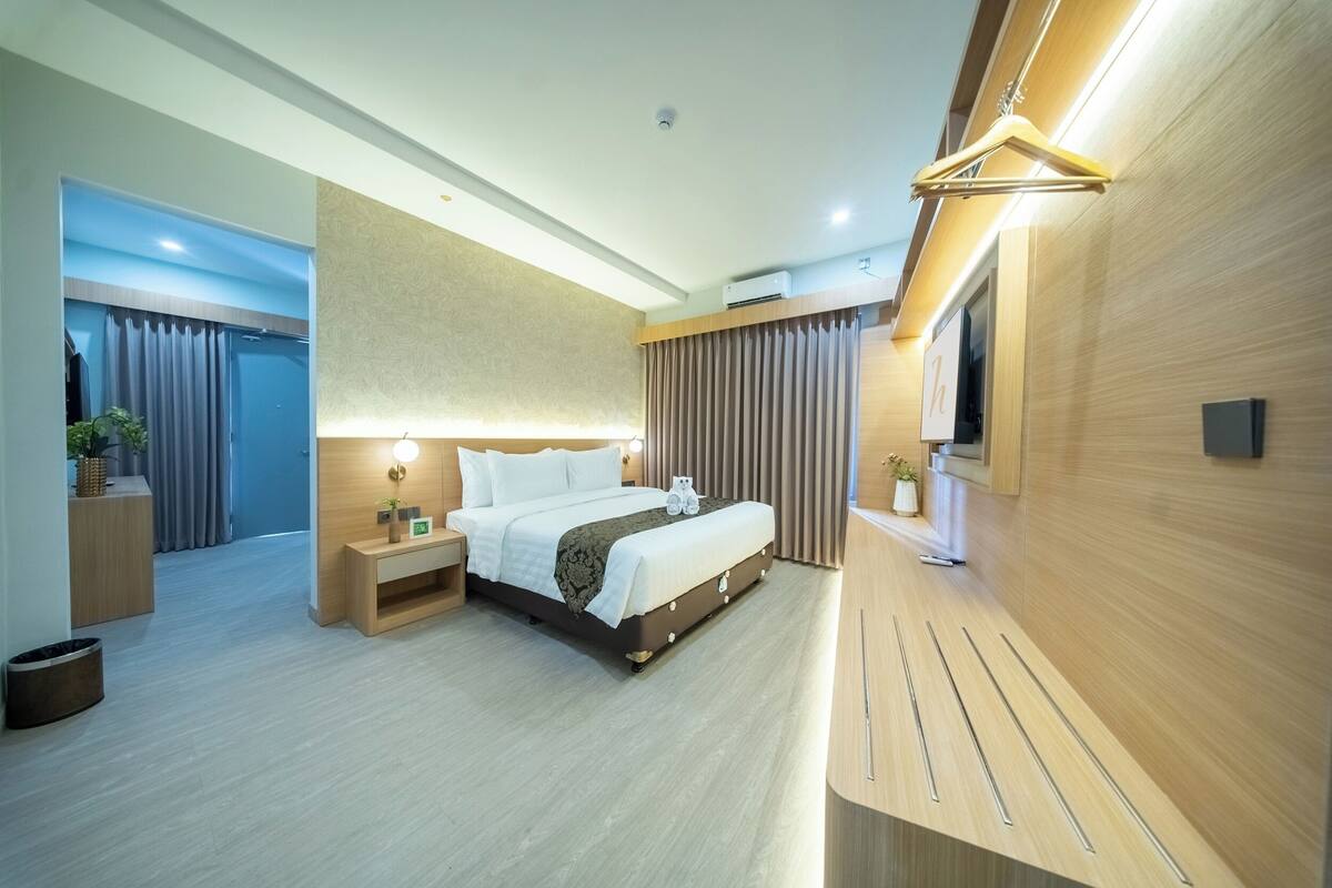 Quarto de hotel moderno com cama, mesa de cabeceira, luminária e televisão na parede.