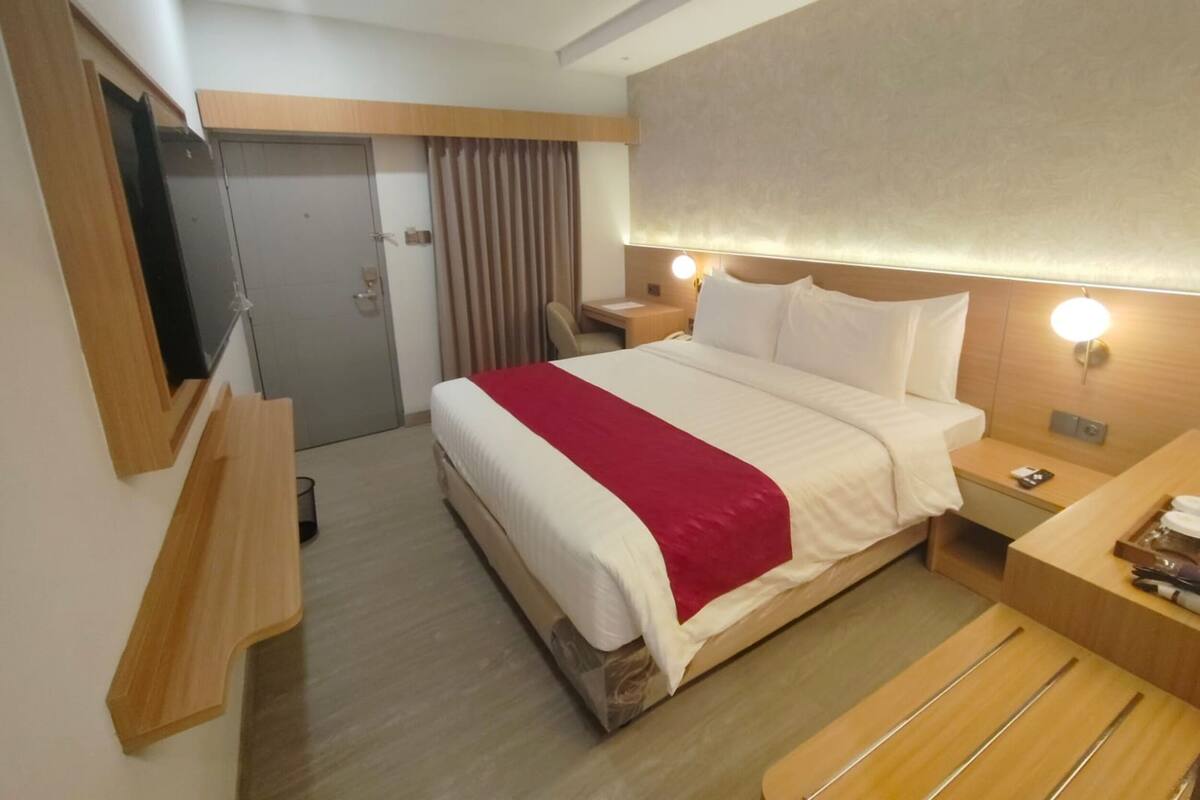 Quarto de hotel com cama, mesas de cabeceira, televisão, cortinas e cabeceira de madeira.