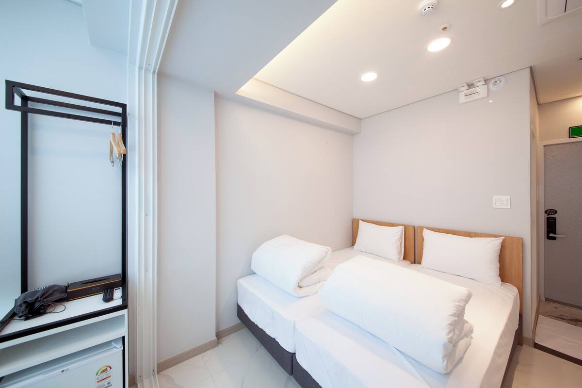 Quarto de hotel com cama, escrivaninha com cadeira, cabideiro e porta.