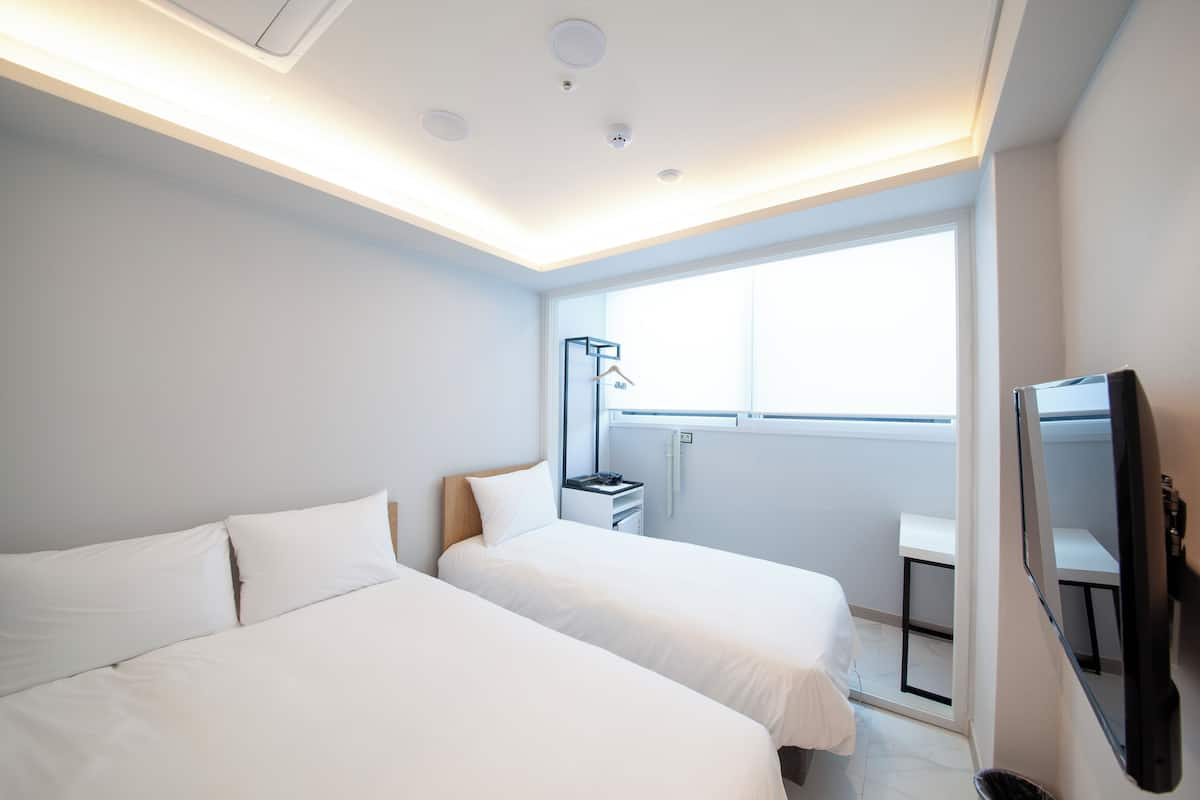 Quarto de hotel moderno com duas camas, televisão, escrivaninha e uma janela grande.