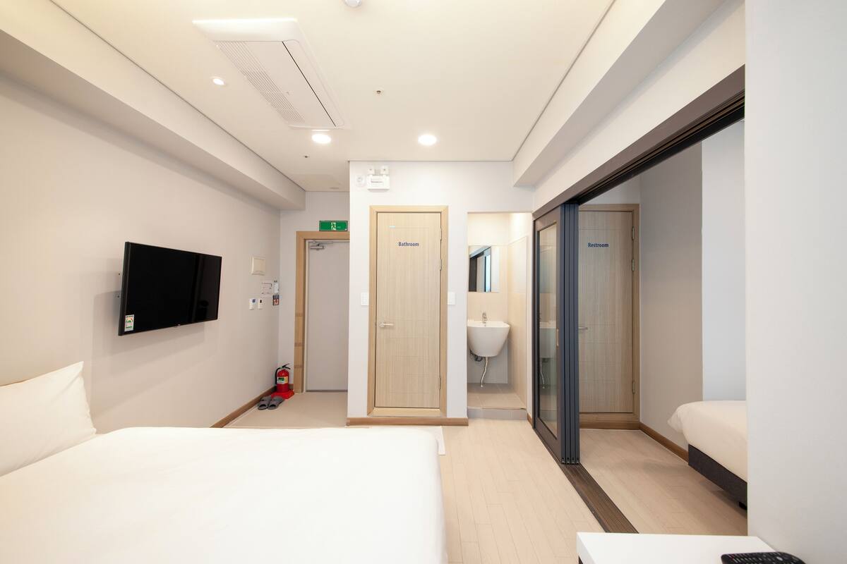 Quarto de hotel com uma cama, televisão na parede, banheiro com pia e vaso sanitário, e porta de vidro deslizante que dá para uma varanda.