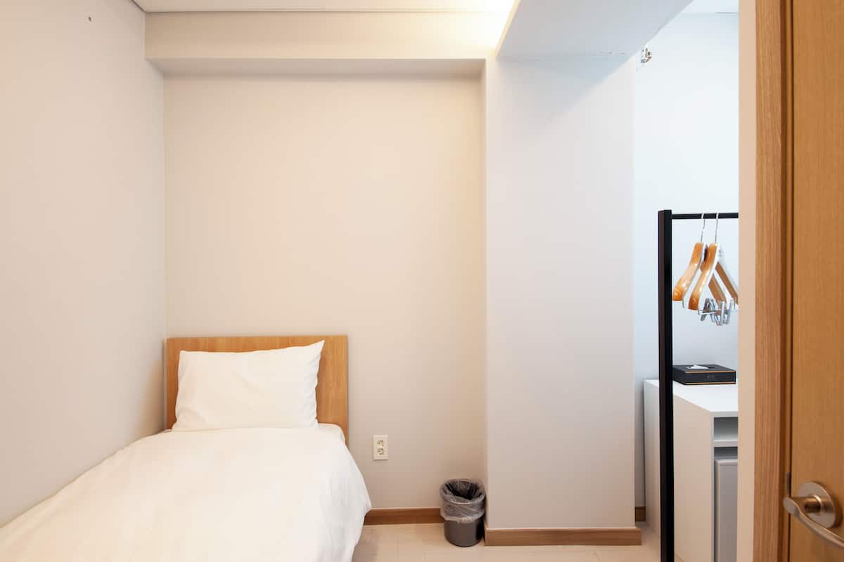 Quarto pequeno e bem arrumado com uma cama de solteiro, cabeceira de madeira, uma pequena lixeira e um guarda-roupa com roupas penduradas.