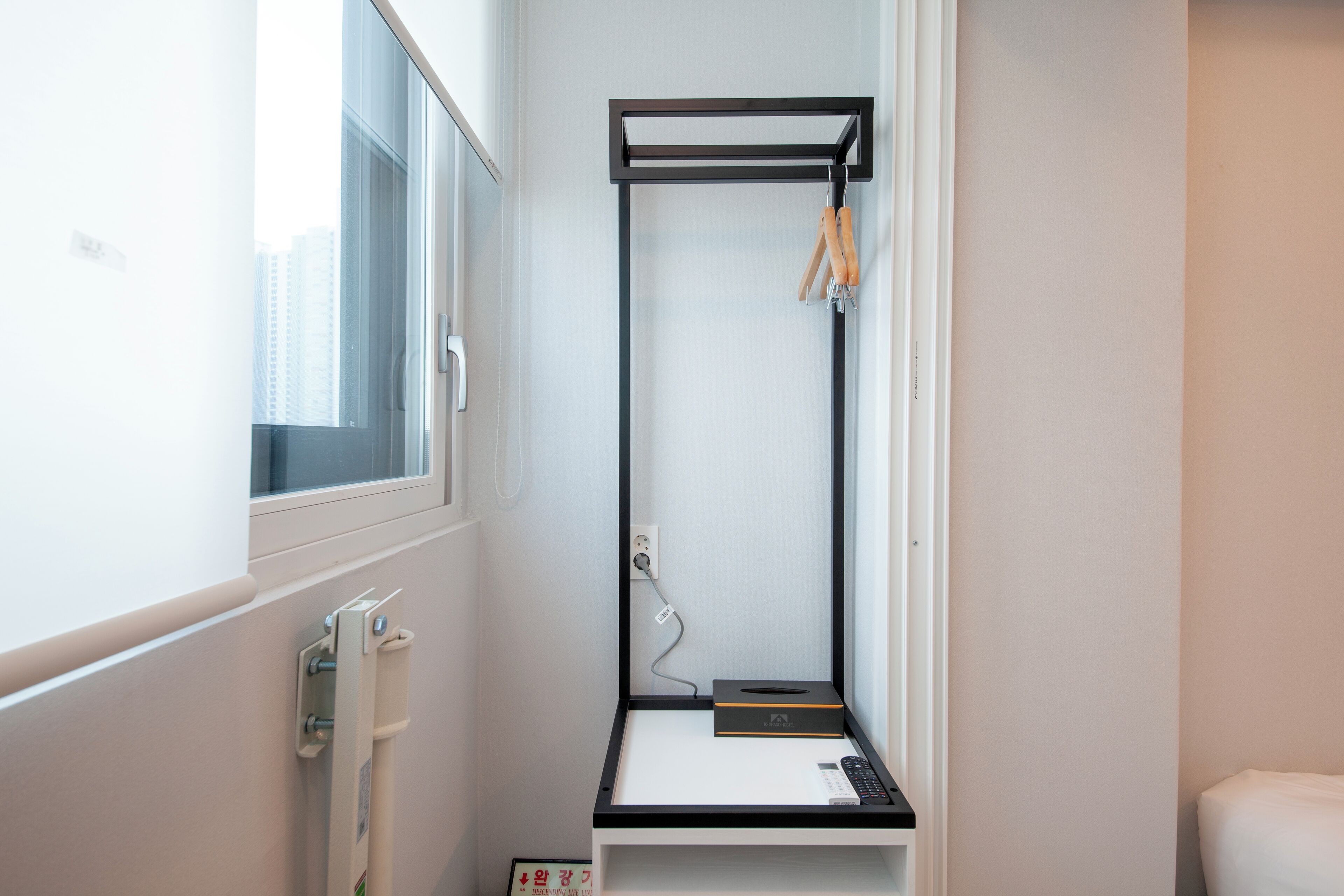 Quarto pequeno com janela, uma prateleira de metal preta com um controle remoto e uma porta branca.