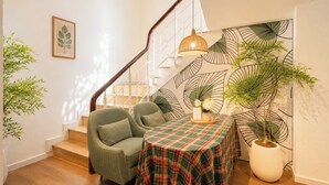 Uma escada com corrimão de madeira, uma poltrona verde, uma mesa coberta por um tecido xadrez e um vaso com planta.