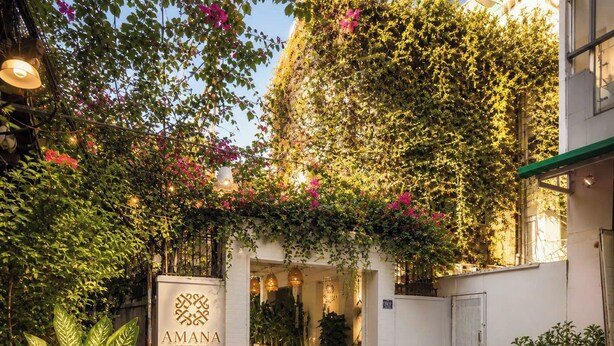 Entrada de um hotel boutique com fachada branca, uma placa indicando 'AMANA BOUTIQUE' e uma área de acesso coberta com plantas em vasos.