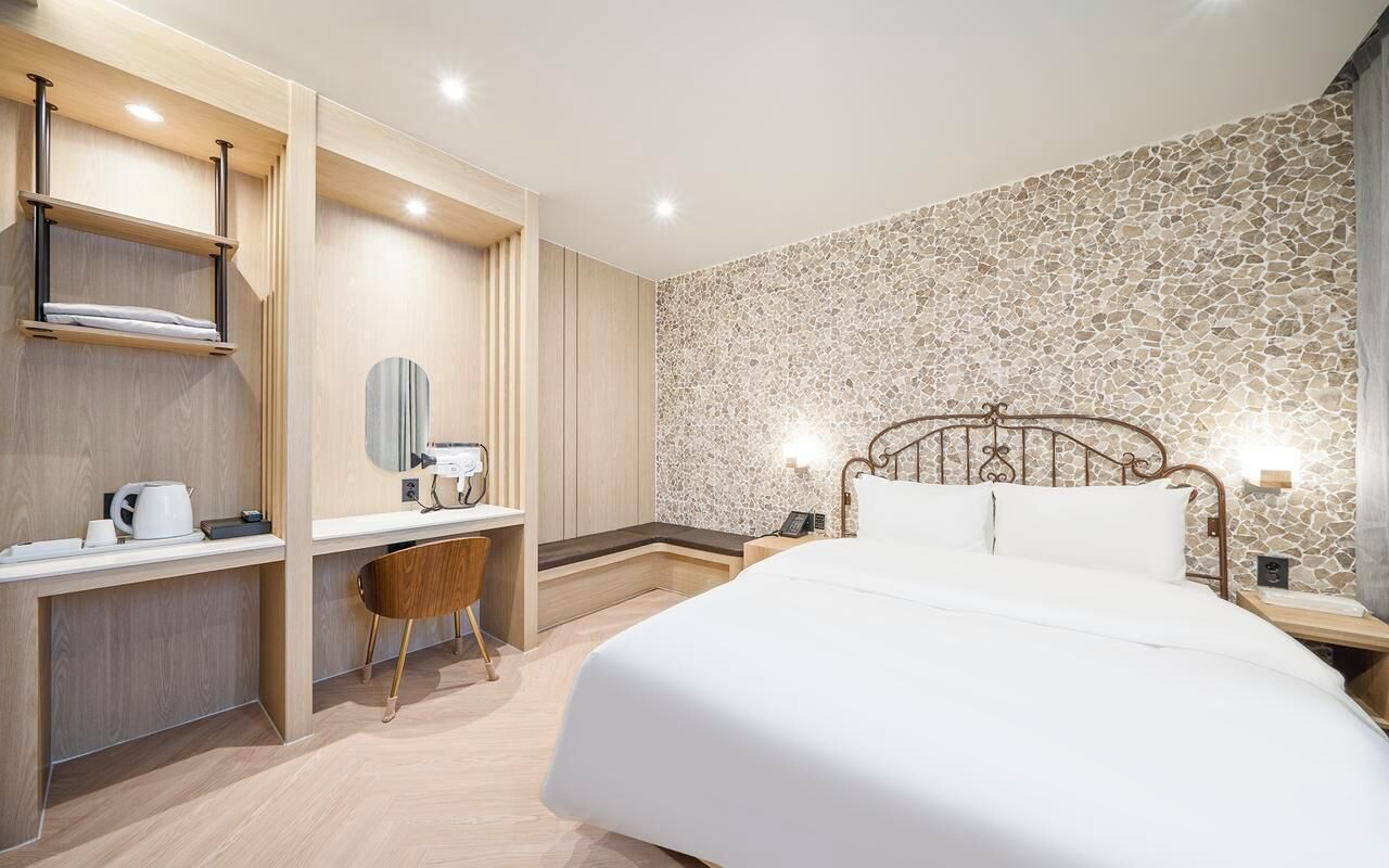 Quarto de hotel com cama, escrivaninha, cadeira, mesa pequena e criado-mudo.