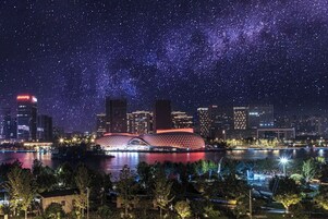 Paisagem urbana noturna com um estádio vermelho iluminado em destaque e um rio ao primeiro plano.