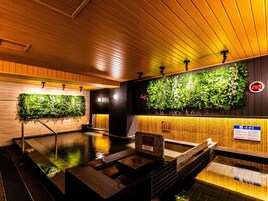 Uma área de spa com teto de madeira, uma planta verde suspensa na parede, um relógio digital e uma bacia de pedra.