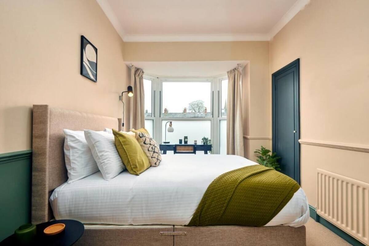 Um quarto com cama, mesa de cabeceira, luminária, janela com cortinas e radiador.