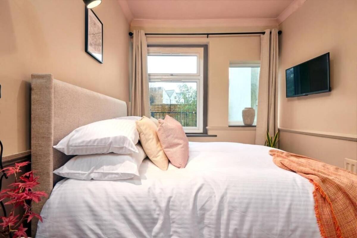 Cama bem arrumada com roupa de cama branca e bege, televisão na parede e janela com cortinas.