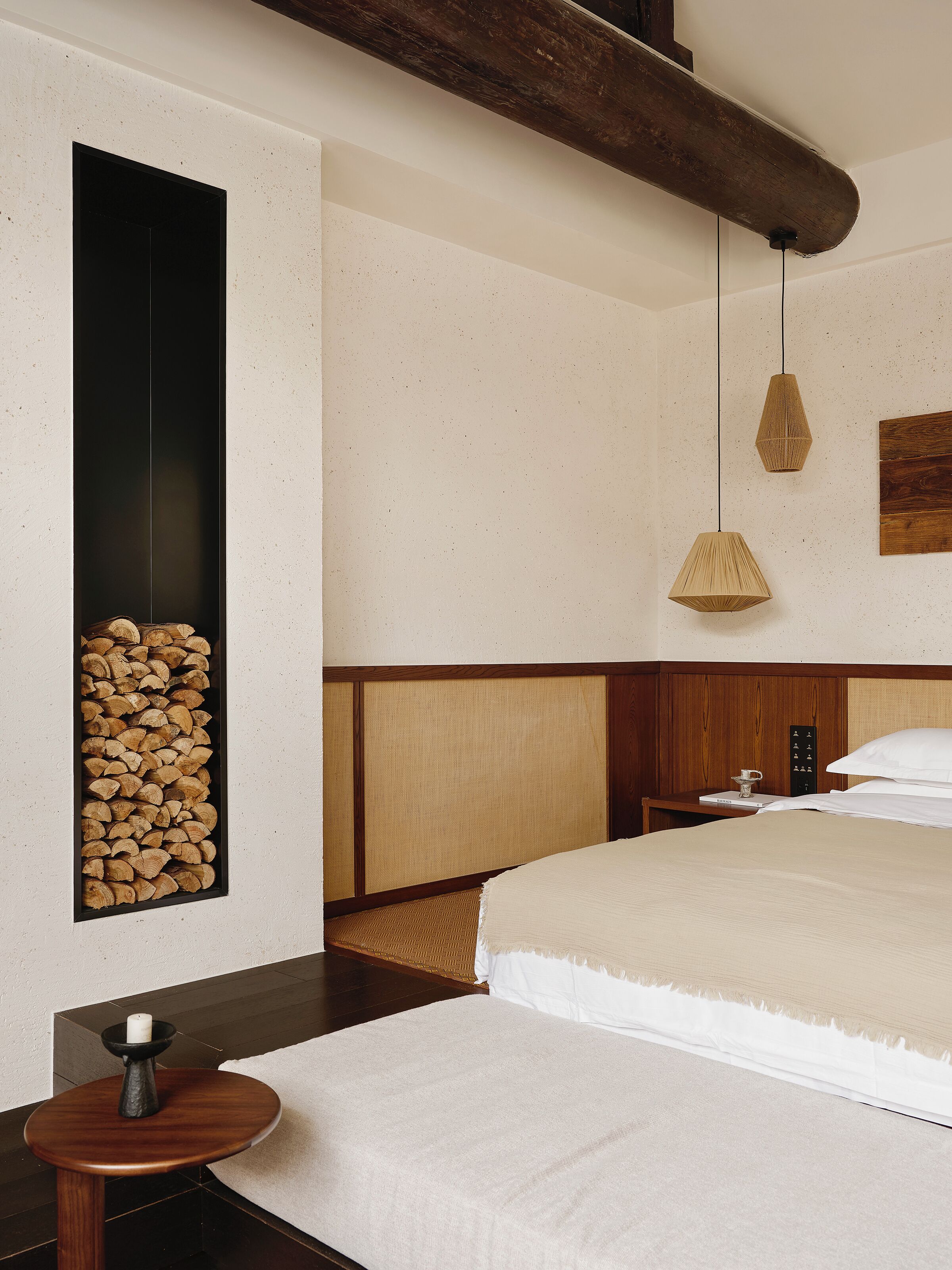 Quarto de hotel com uma cama grande, cabeceira de madeira, prateleira na parede com toras, mesa de cabeceira com uma vela e luminária pendente.