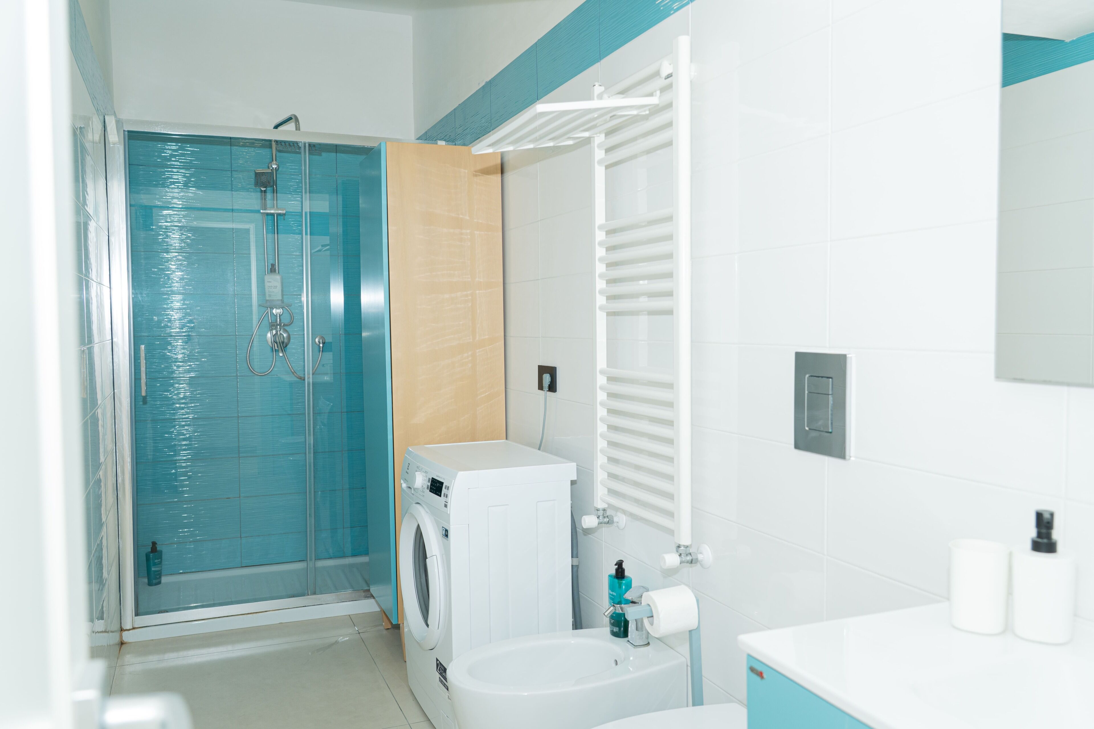 Uma casa de banho moderna com cabine de chuveiro em vidro, máquina de lavar roupa embutida e lavatório branco com barra de toalhas.
