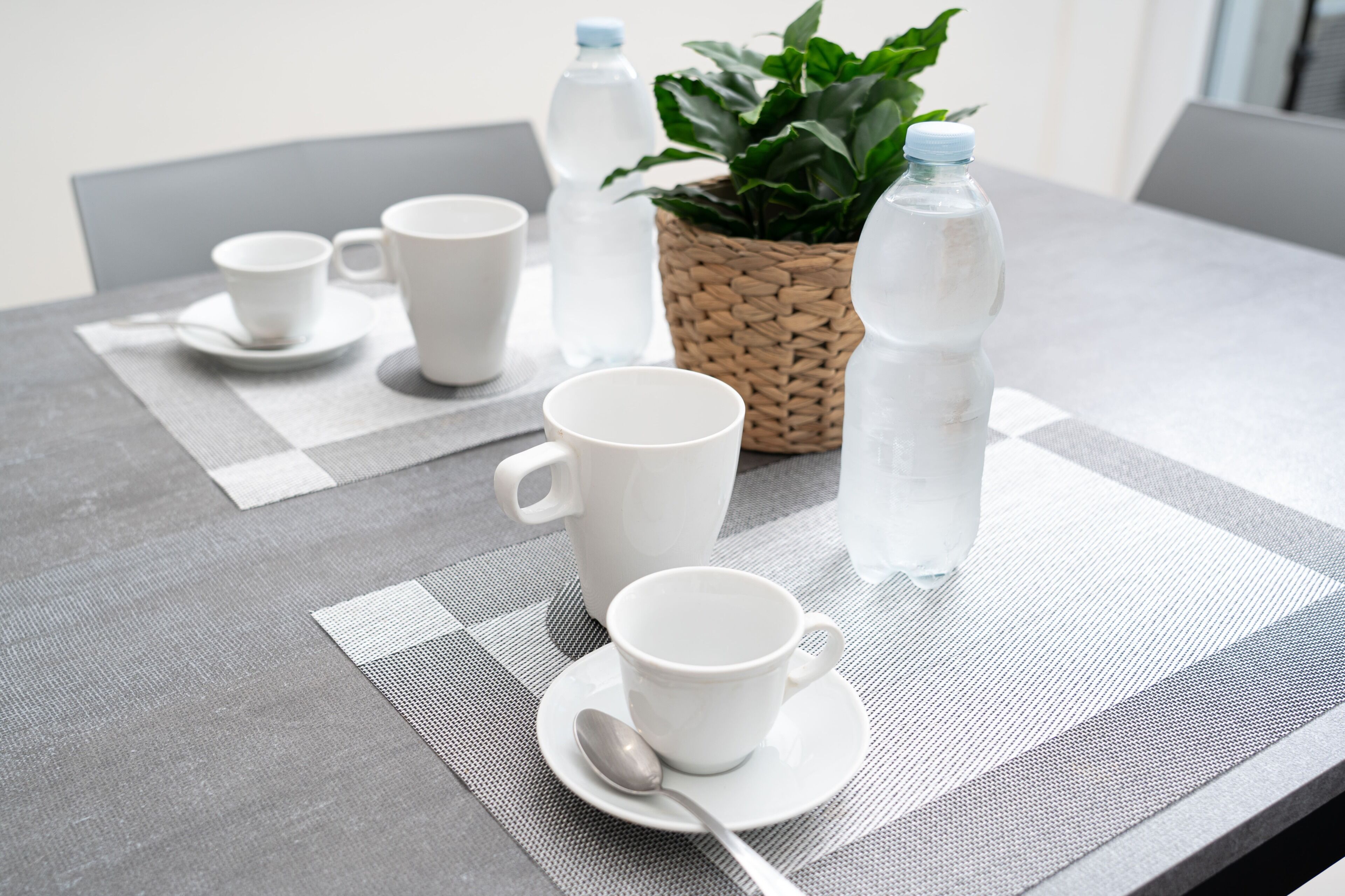 Uma mesa com xícaras, pires e uma colher, além de duas garrafas de água e uma cesta de vime com uma planta.