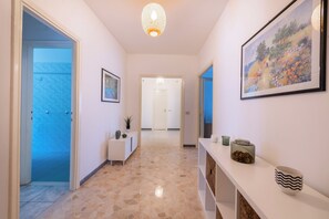 Um corredor com piso ladrilhado, um console branco com objetos decorativos, um quadro na parede e uma porta que dá para um quarto com paredes azulejadas.