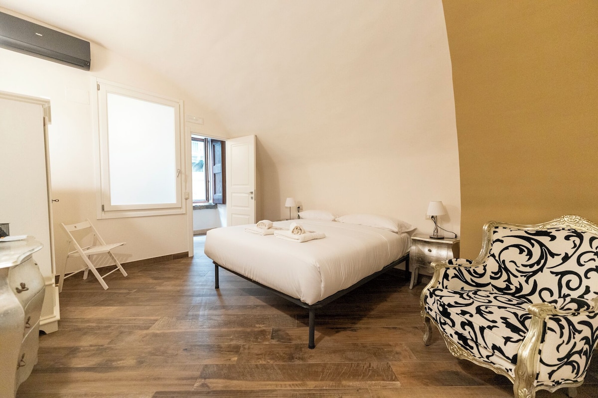 Quarto com cama, mesas de cabeceira, uma cadeira e janela.