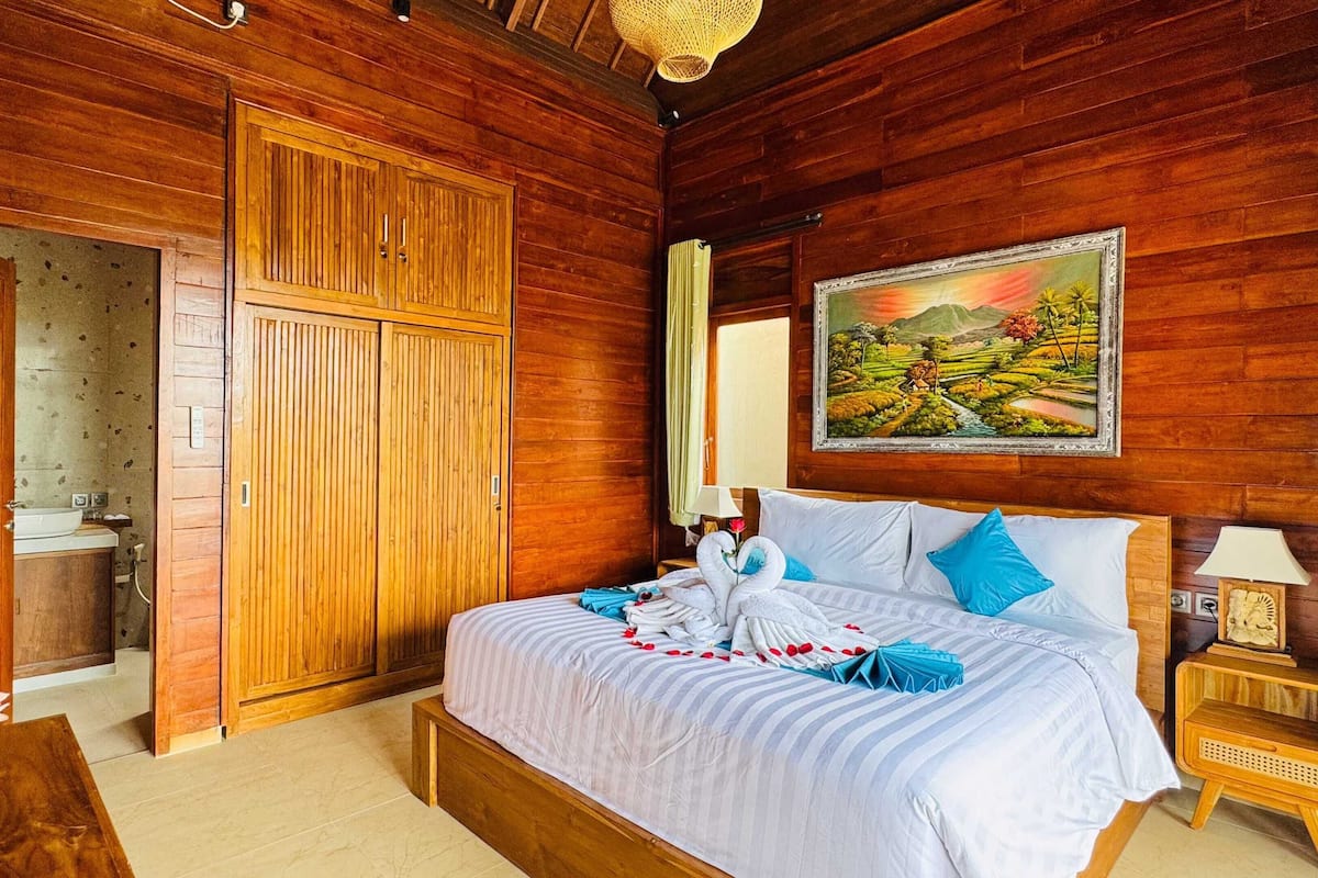Quarto de madeira com cama, um quadro, um guarda-roupa de madeira e um banheiro visível por uma porta aberta.