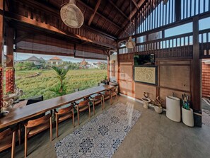 Uma sala de jantar com uma mesa de madeira comprida, cadeiras, um tapete estampado e vista para um campo verde e casas.