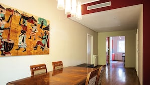 Uma sala de jantar com mesa e cadeiras de madeira, um quadro decorativo grande na parede e um corredor que dá acesso a outros aposentos.