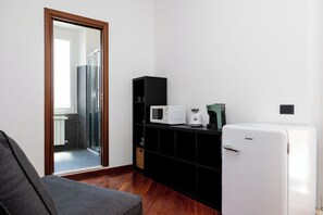 Quarto compacto com porta de vidro deslizante para área de chuveiro, geladeira branca, micro-ondas e estante preta.