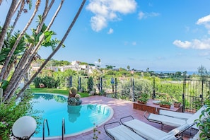 Piscina com espreguiçadeiras, rodeada por vegetação e vista para o mar.