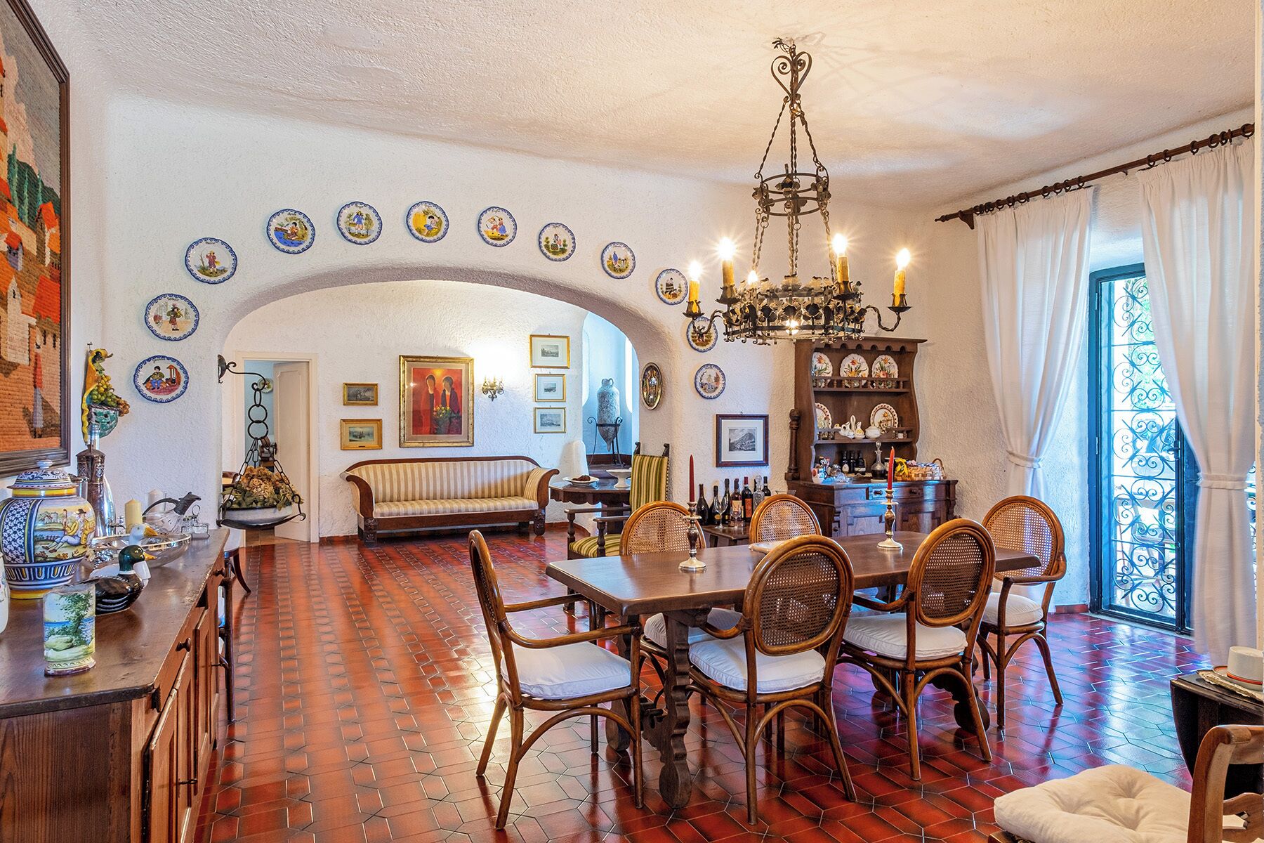 Uma área de refeições com mesa de madeira e cadeiras de vime, um candelabro e uma parede decorada com pratos.