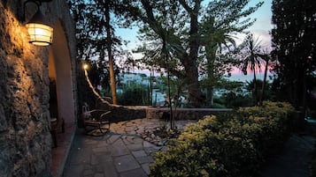 Uma trilha de pedra leva a uma área de estar com uma cadeira, um candeeiro e vista para uma cidade costeira ao entardecer.