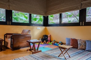Quarto com cadeira de madeira, pequena mesa, sofá e um tabuleiro de xadrez. Janelas oferecem vista para o verde lá fora.