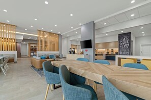 Um hall moderno de hotel com uma longa mesa de jantar, cadeiras azuis, uma área de estar com sofás, uma cozinha e uma grande televisão.