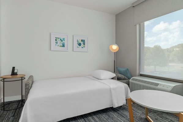 Quarto de hotel com cama, mesa redonda, luminária de pé e duas obras de arte emolduradas na parede.
