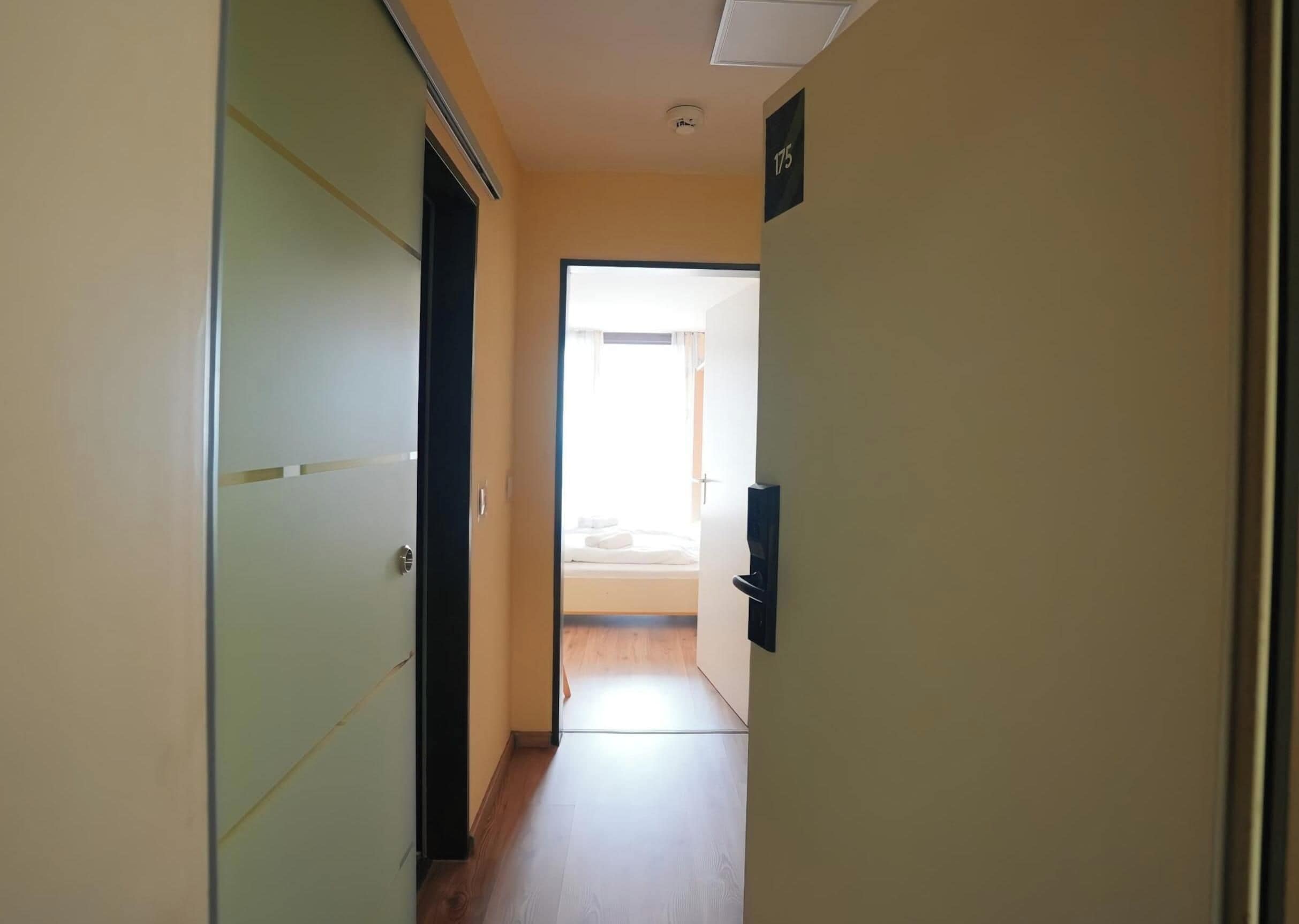 Um corredor estreito com uma porta que dá para um quarto, onde se vê uma cama e uma janela.