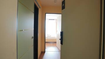 Um corredor estreito com uma porta que dá para um quarto, onde se vê uma cama e uma janela.