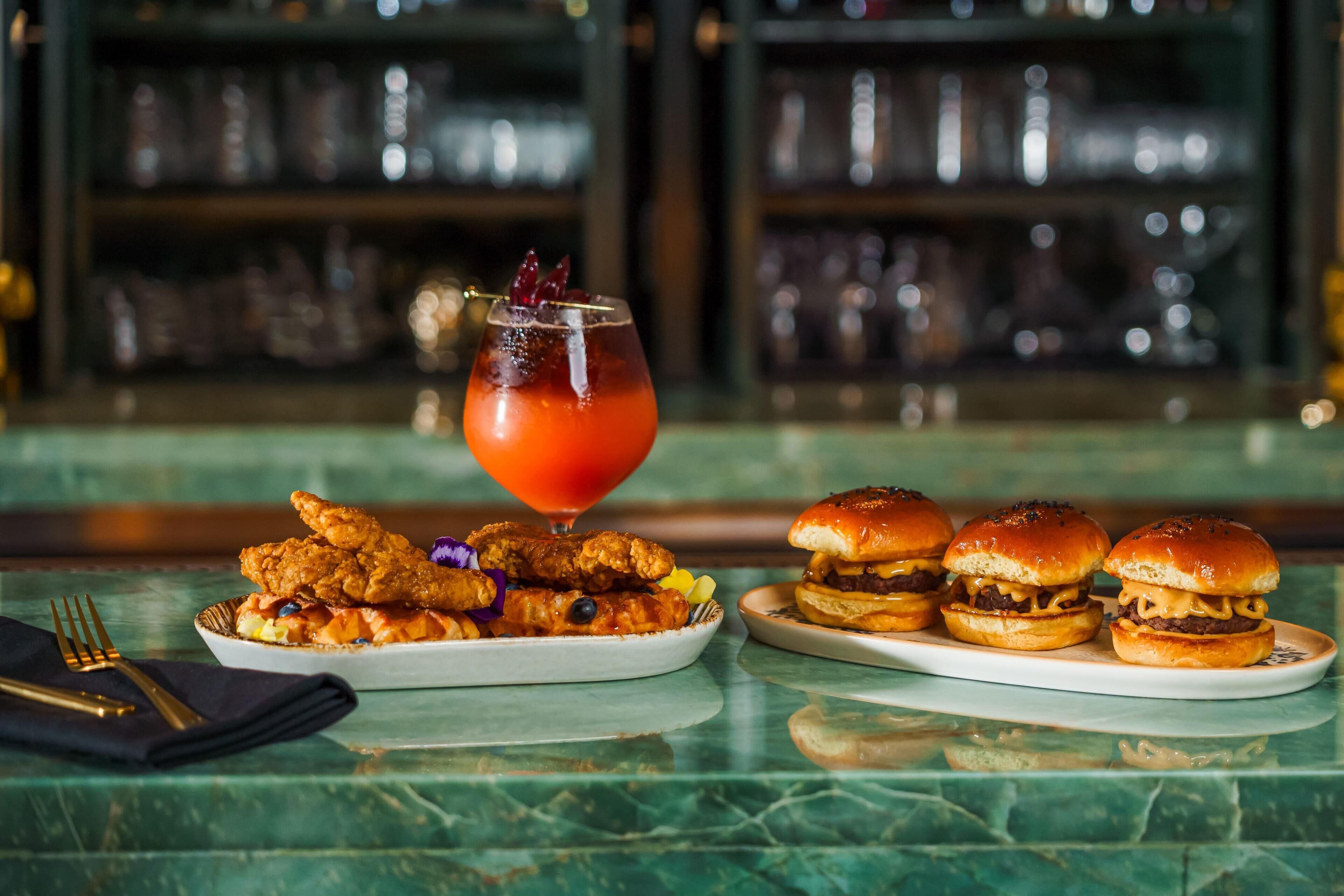 Um balcão de bar com um prato de frango frito, um copo de bebida vermelha e hambúrgueres sobre uma mesa de mármore.